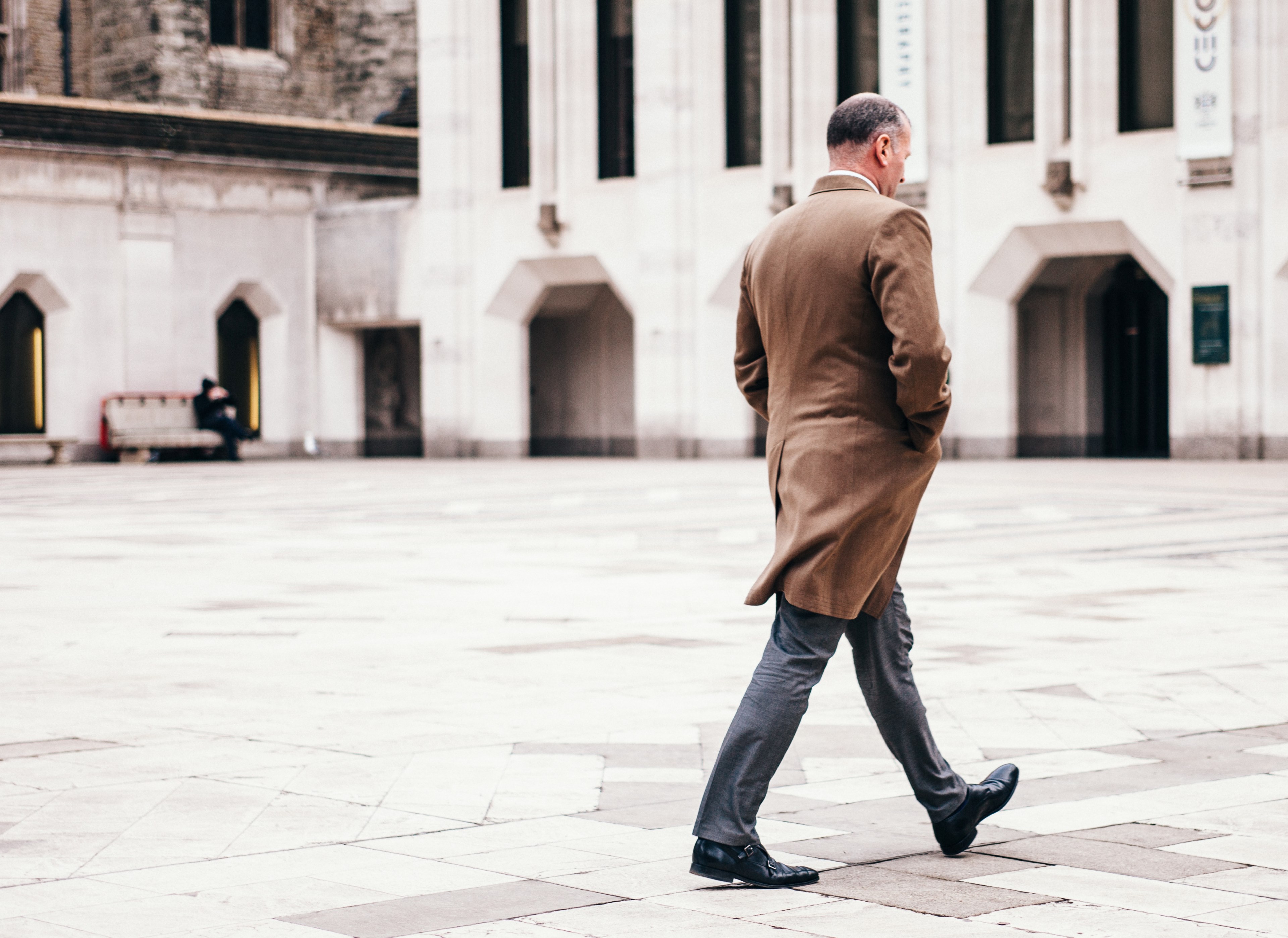 a man in a brown coat walking across an urban plaza, youll never walk ...