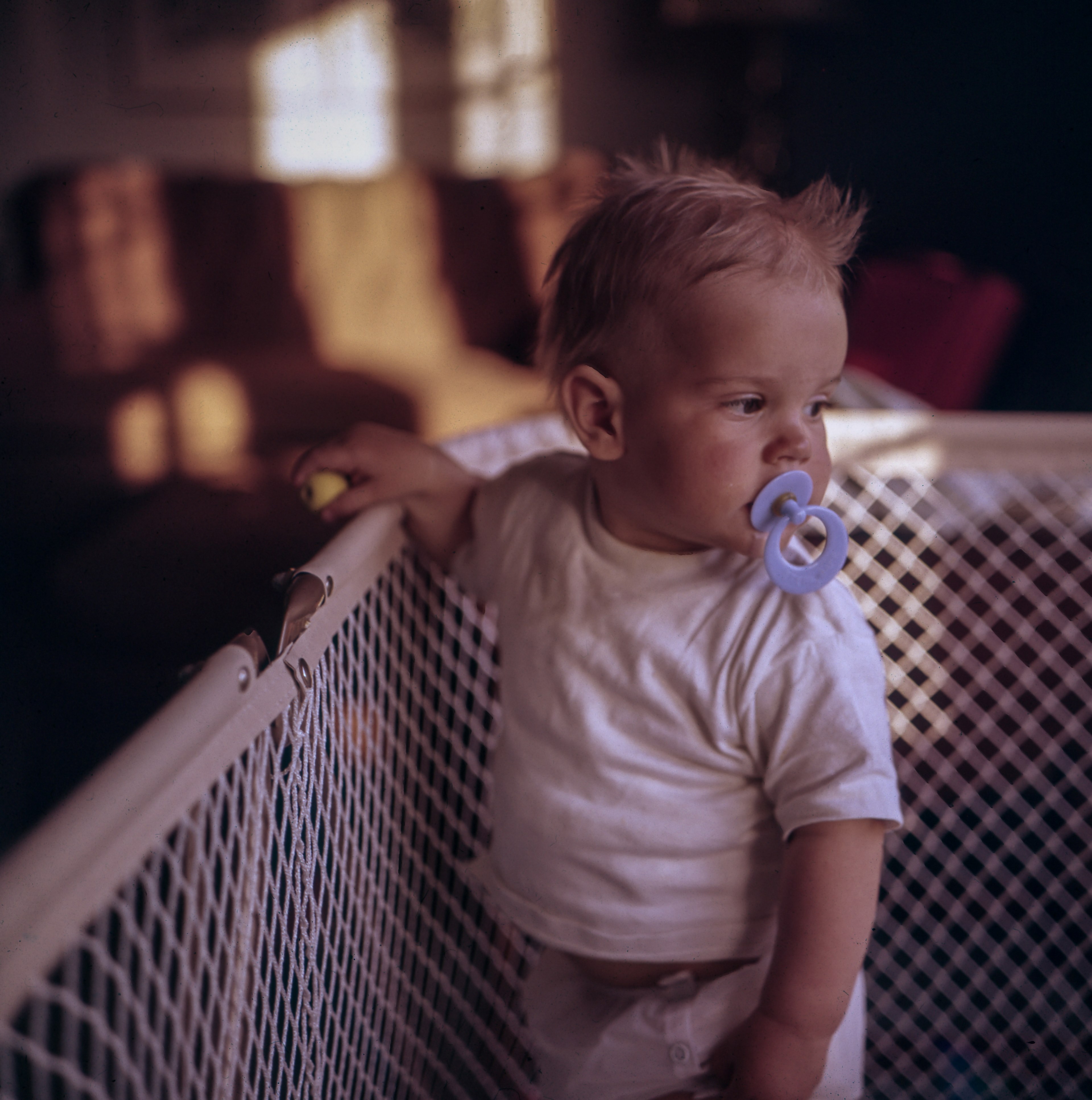a baby with a pacifier standing up in a white crib in a dim room ...